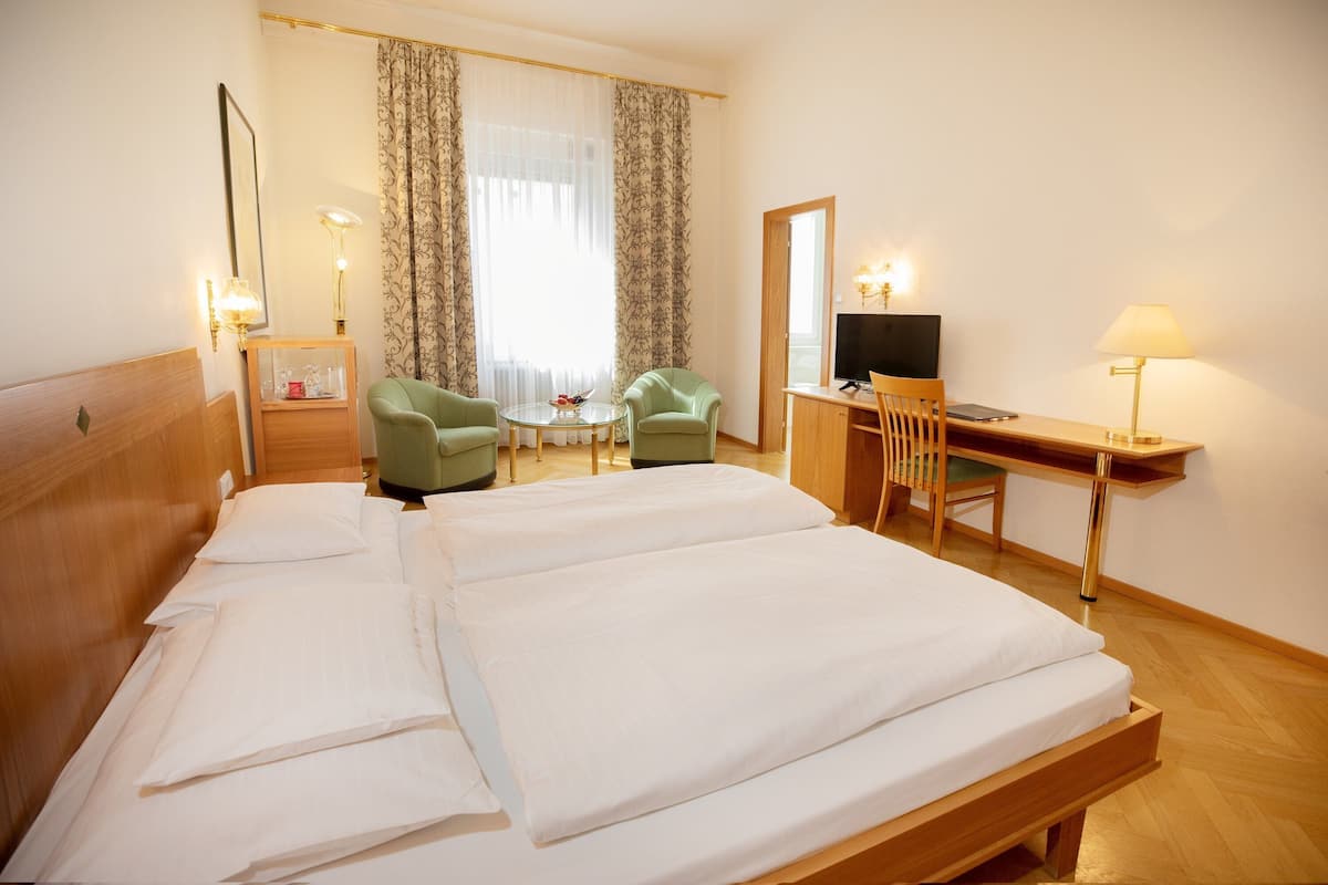 Quarto de hotel com uma cama grande, duas poltronas, uma mesa redonda pequena, uma escrivaninha com cadeira, uma televisão e um abajur.