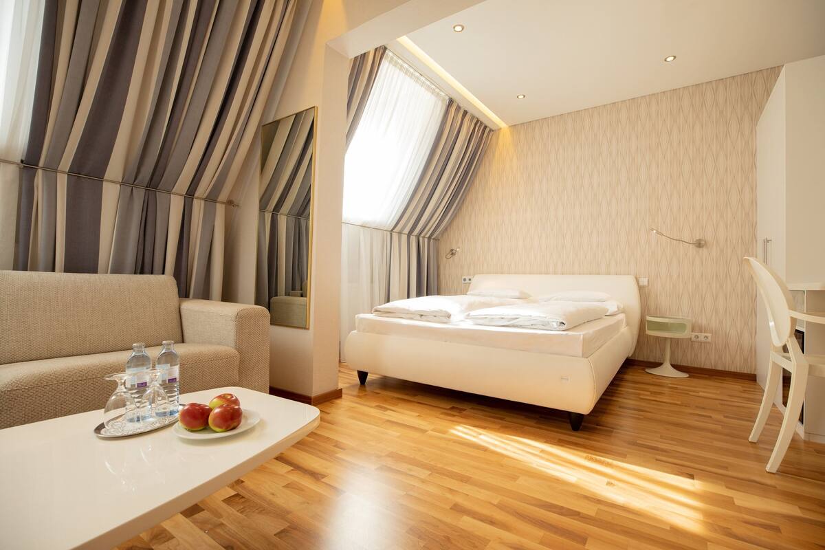 Quarto de hotel moderno com cama, sofá, mesa de jantar com maçãs e garrafas de água, e uma pequena escrivaninha.