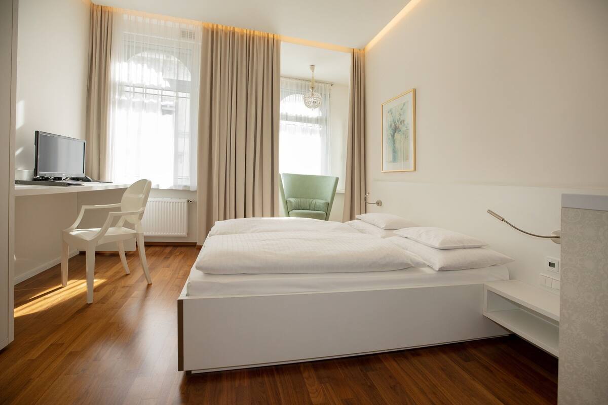 Quarto de hotel moderno com cama, escrivaninha com computador, cadeira e janela com cortinas.