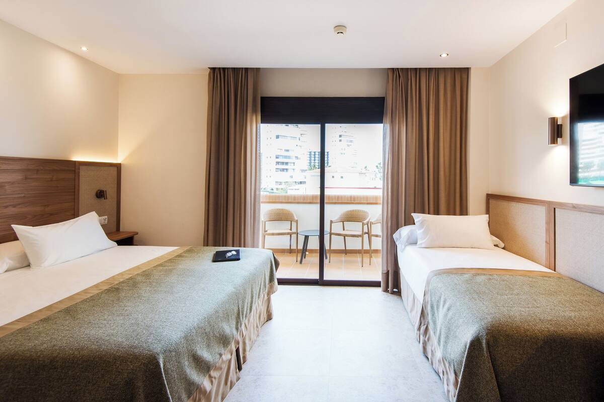 Quarto de hotel com duas camas, varanda com cadeiras e vista para edifícios.