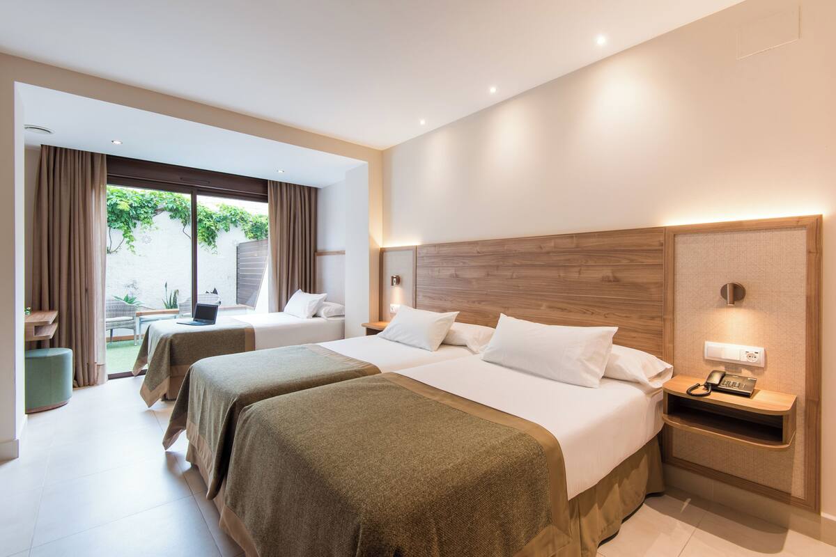 Quarto de hotel com duas camas, uma escrivaninha, uma cadeira, uma mesinha, um telefone e uma janela com cortinas.