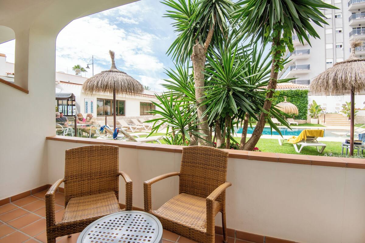 Varanda com cadeiras de vime e uma mesa redonda, com vista para uma área de piscina com guarda-sóis e palmeiras.