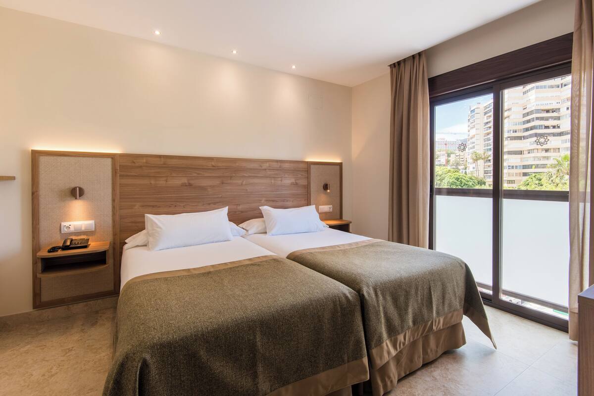 Quarto de hotel com duas camas, cabeceira de madeira, luminárias de cabeceira, mesa de cabeceira e uma janela grande com vista para edifícios.