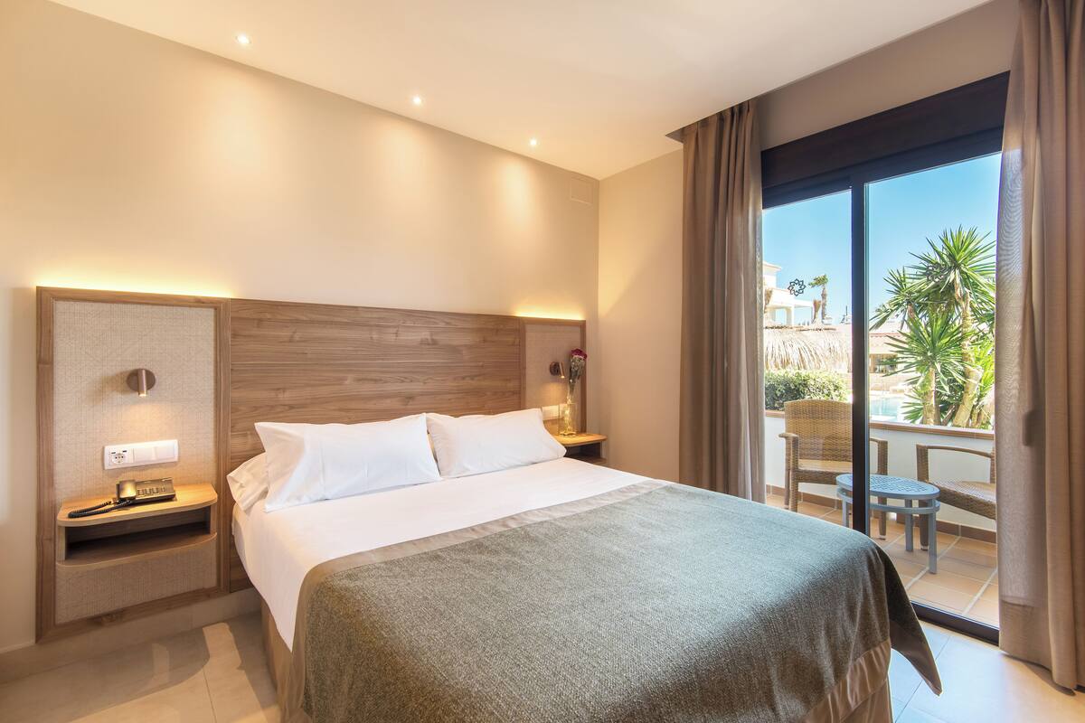 Quarto de hotel com uma cama grande, mesa de cabeceira, luminária e vista para a varanda externa.