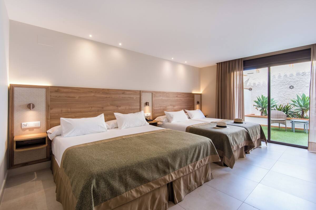 Quarto de hotel com duas camas, cabeceira de madeira, uma mesinha e vista para um jardim através de uma porta de vidro.