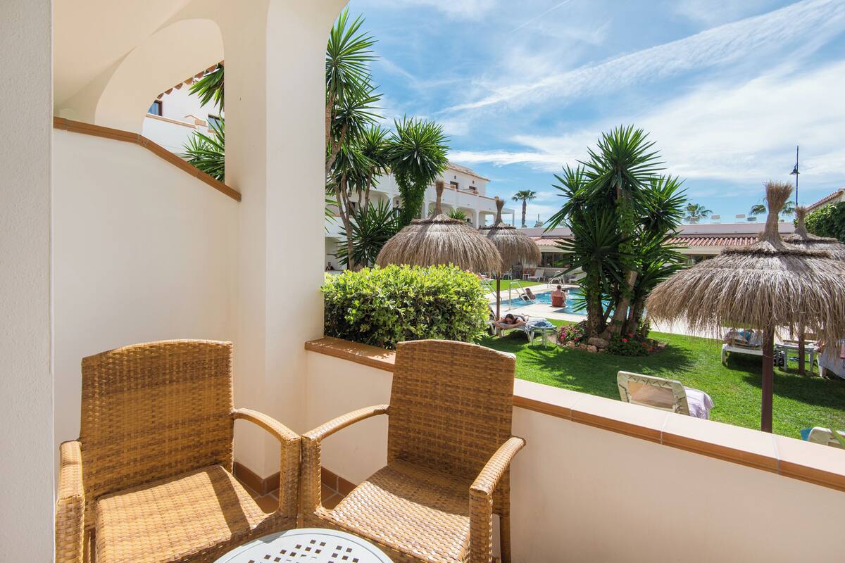 Uma varanda com cadeiras de vime, uma mesa pequena e vista para uma área de piscina com palmeiras e guarda-sóis de palha.