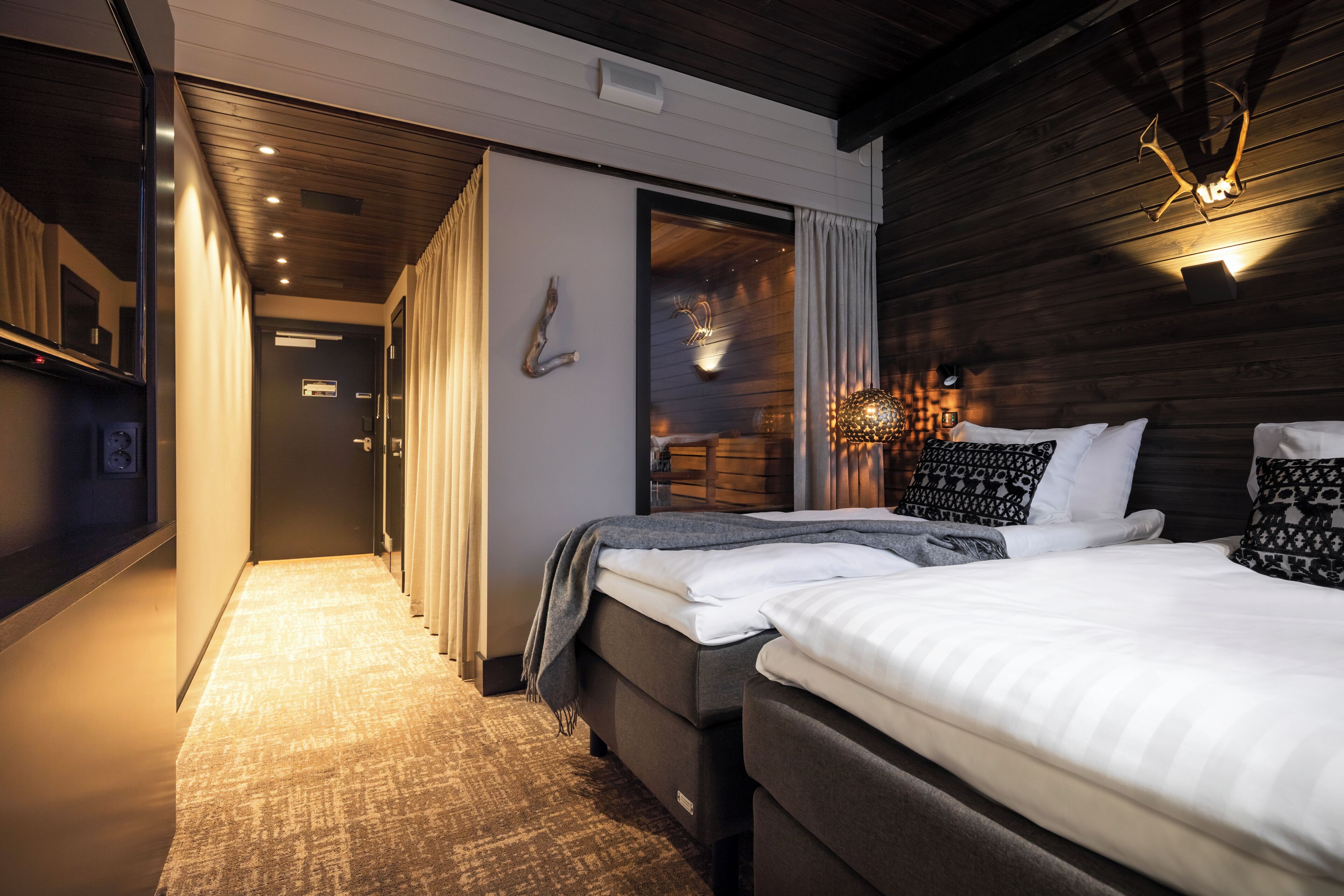 Quarto de hotel moderno com teto de madeira, uma cama grande com almofadas estampadas, mesa de cabeceira e luminária fixada na parede.