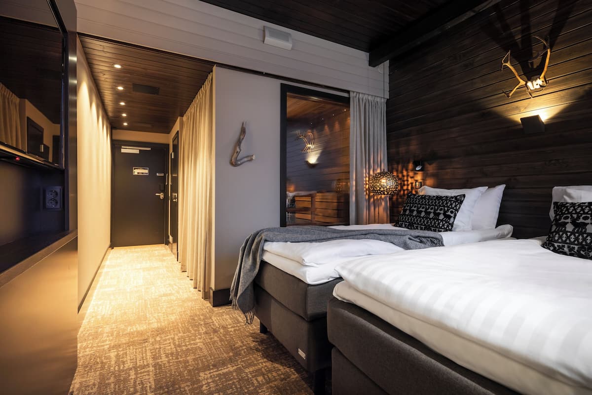 Quarto de hotel moderno com teto de madeira, uma cama grande com almofadas estampadas, mesa de cabeceira e luminária fixada na parede.