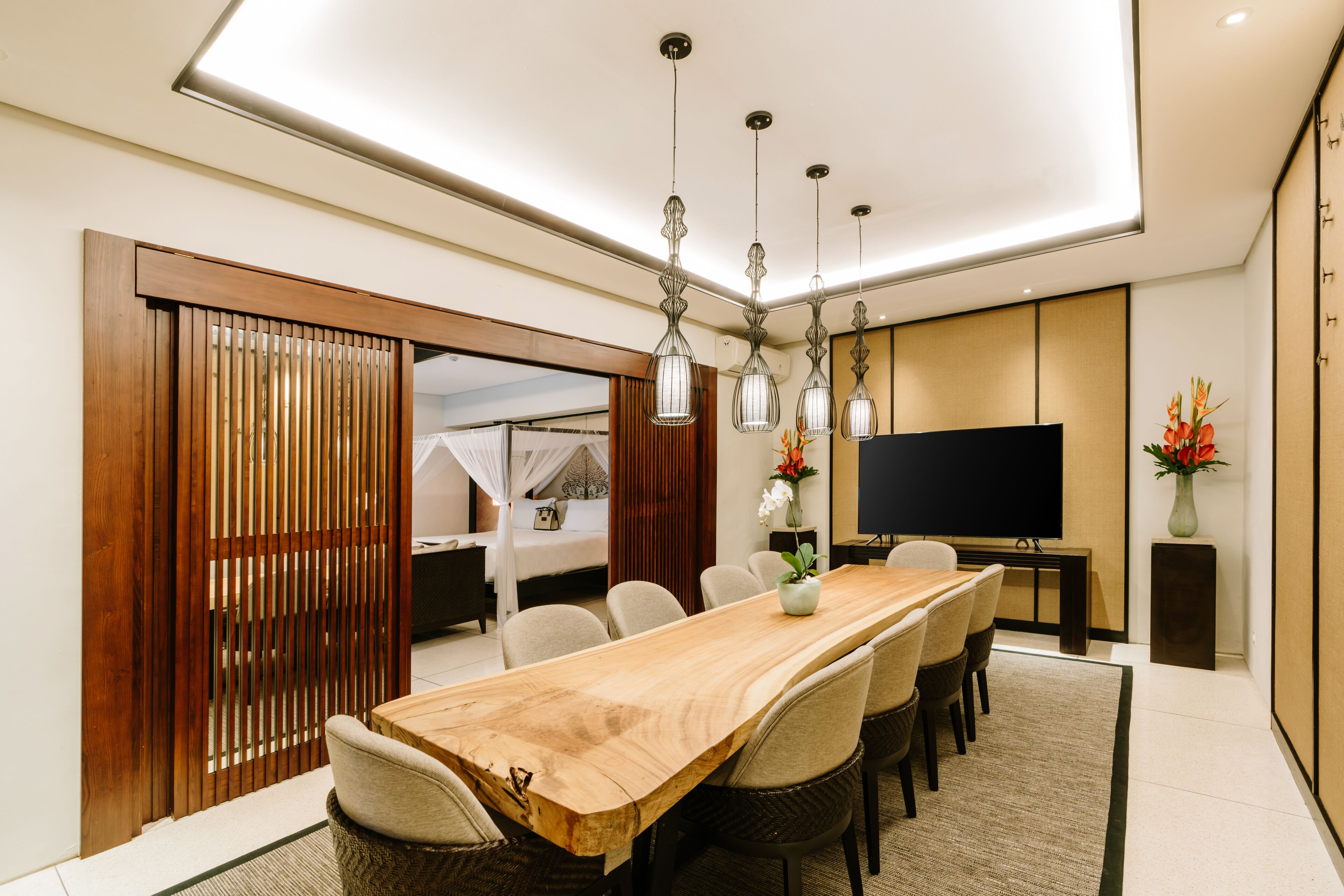 Uma sala de jantar moderna com uma longa mesa de madeira, várias cadeiras, luminárias pendentes, uma televisão de tela plana e um vaso decorativo com flores.