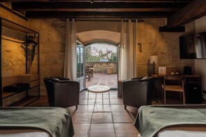 Quarto com cama, duas cadeiras, uma mesinha, televisão e vista para um pátio externo.