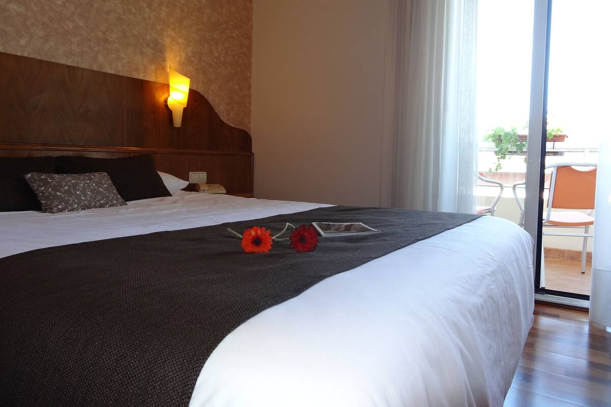 Quarto de hotel com cama grande, mesa de cabeceira, luminária na parede e vista para a varanda externa.
