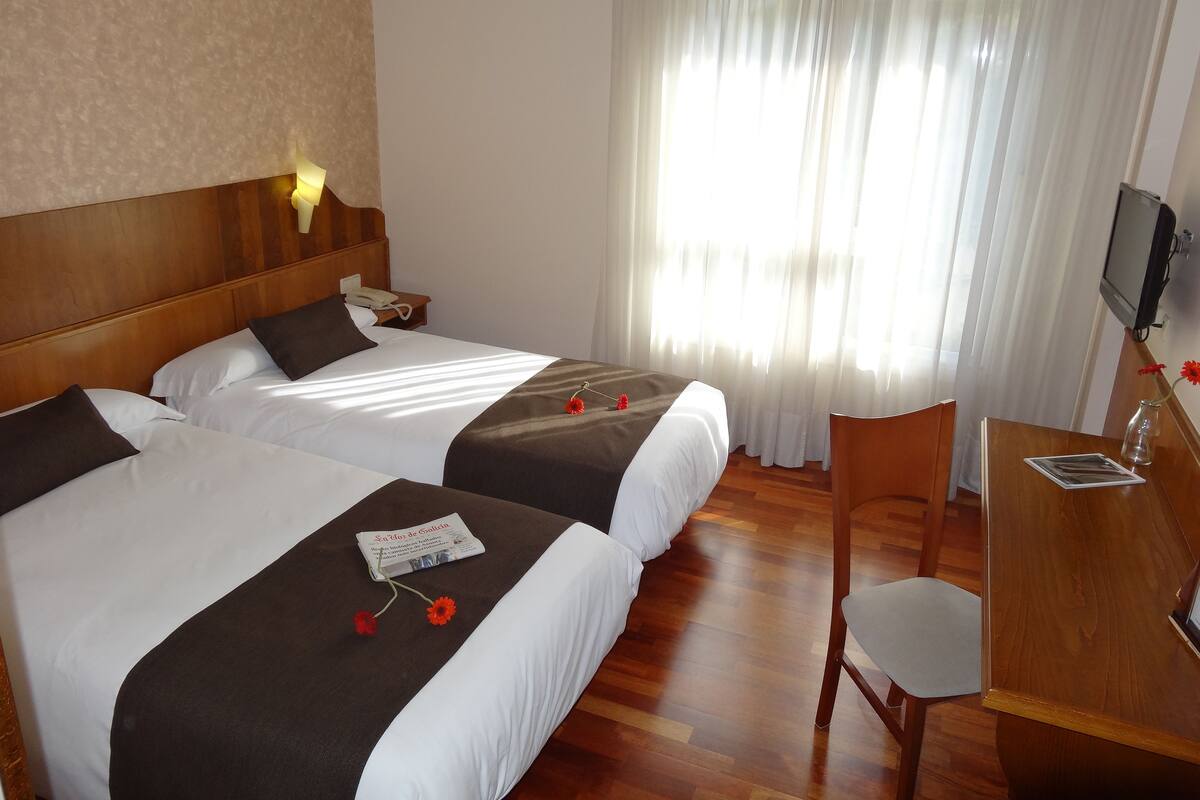 Quarto de hotel com duas camas, uma escrivaninha, uma cadeira, uma televisão e uma janela com cortinas.