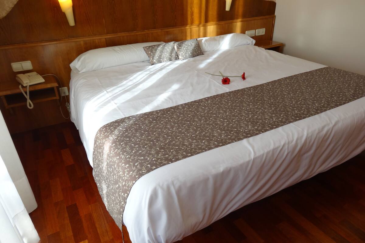 Quarto de hotel com cabeceira de madeira, telefone na mesa de cabeceira e cama com roupa de cama branca e um corredor estampado.