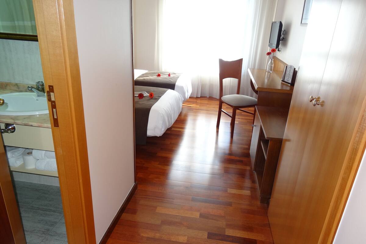 Quarto de hotel com piso de madeira, uma cama com roupa de cama branca, uma cadeira de madeira, uma escrivaninha com um vaso e uma televisão na parede.