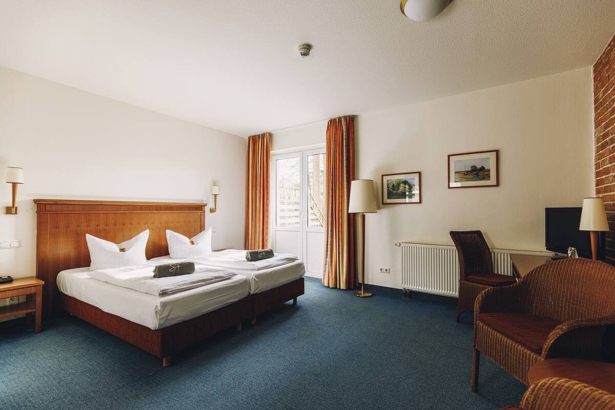 Quarto de hotel com cama de casal, uma escrivaninha com cadeira, televisão e janela com cortinas.