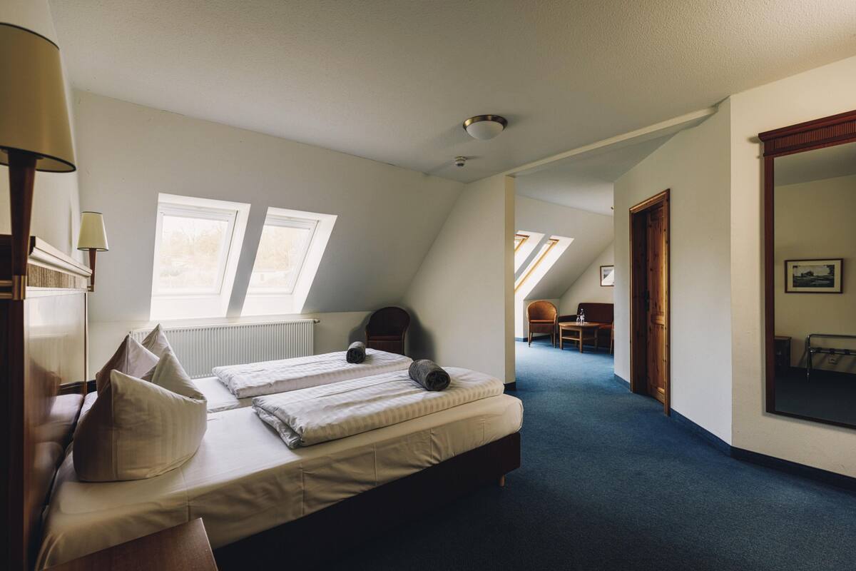 Quarto com duas camas, uma escrivaninha e uma cadeira. Há janelas e uma porta que dá para outro cômodo.