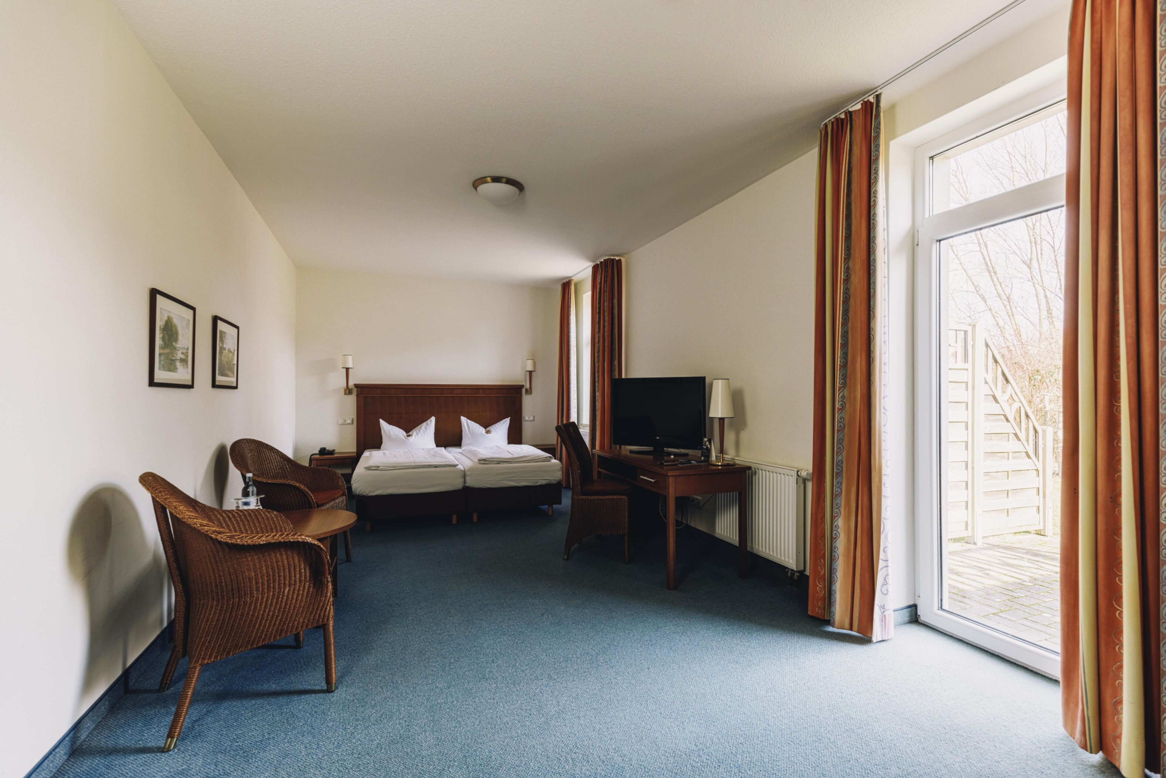 Quarto de hotel com cama, escrivaninha com televisão, cadeira e janela com cortinas.