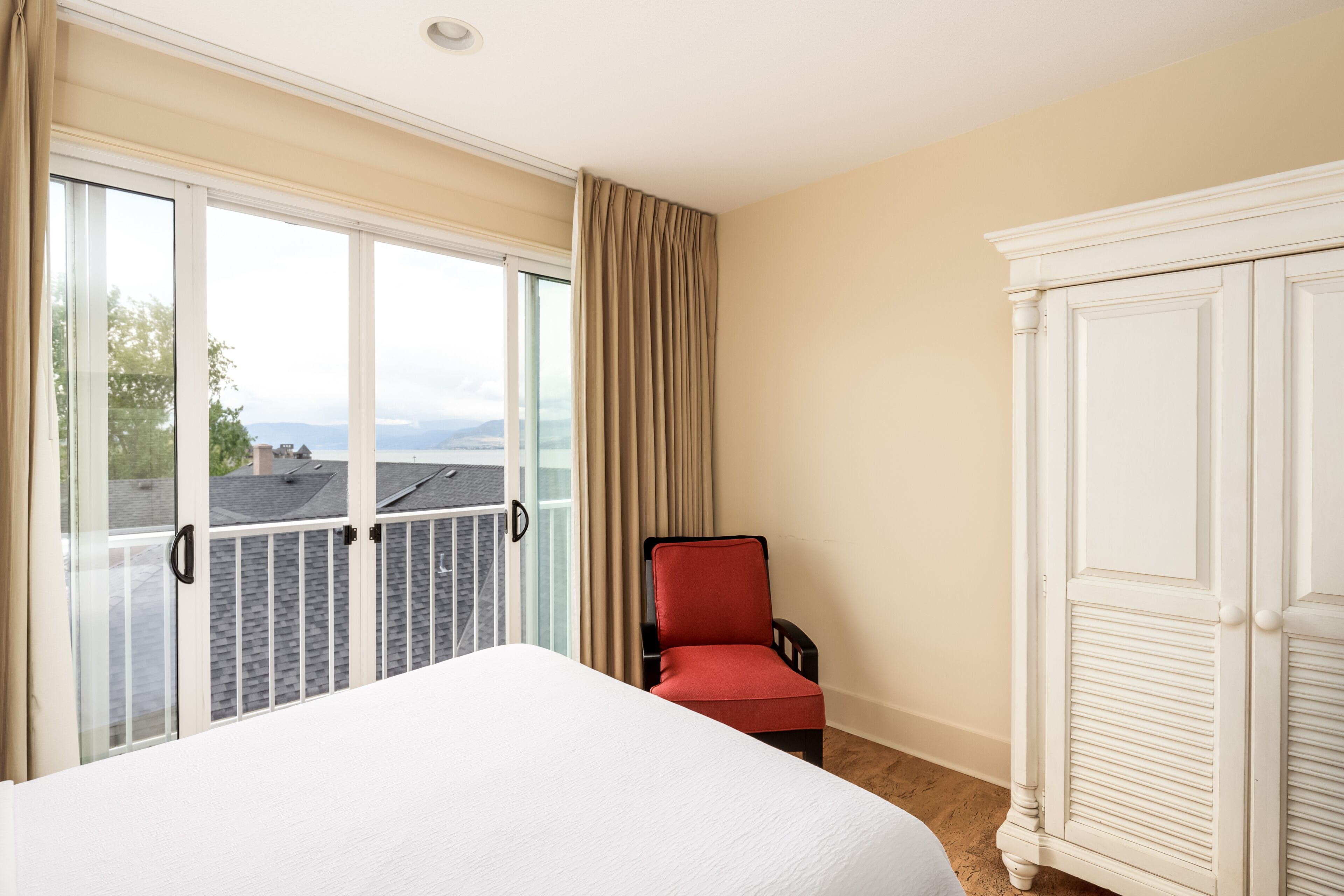 Quarto de hotel com uma cama grande, uma cadeira vermelha, um guarda-roupa e varanda com vista para a paisagem urbana.