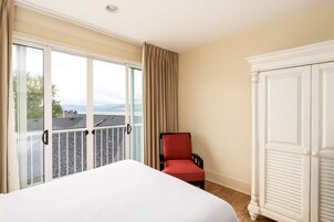 Quarto de hotel com uma cama grande, uma cadeira vermelha, um guarda-roupa e varanda com vista para a paisagem urbana.