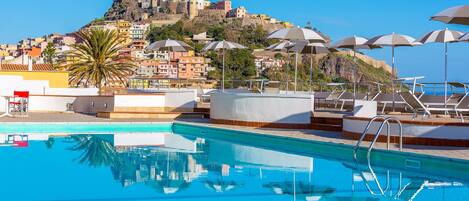 Uma piscina com espreguiçadeiras e guarda-sóis, céu azul límpido e uma colina com edifícios ao fundo.
