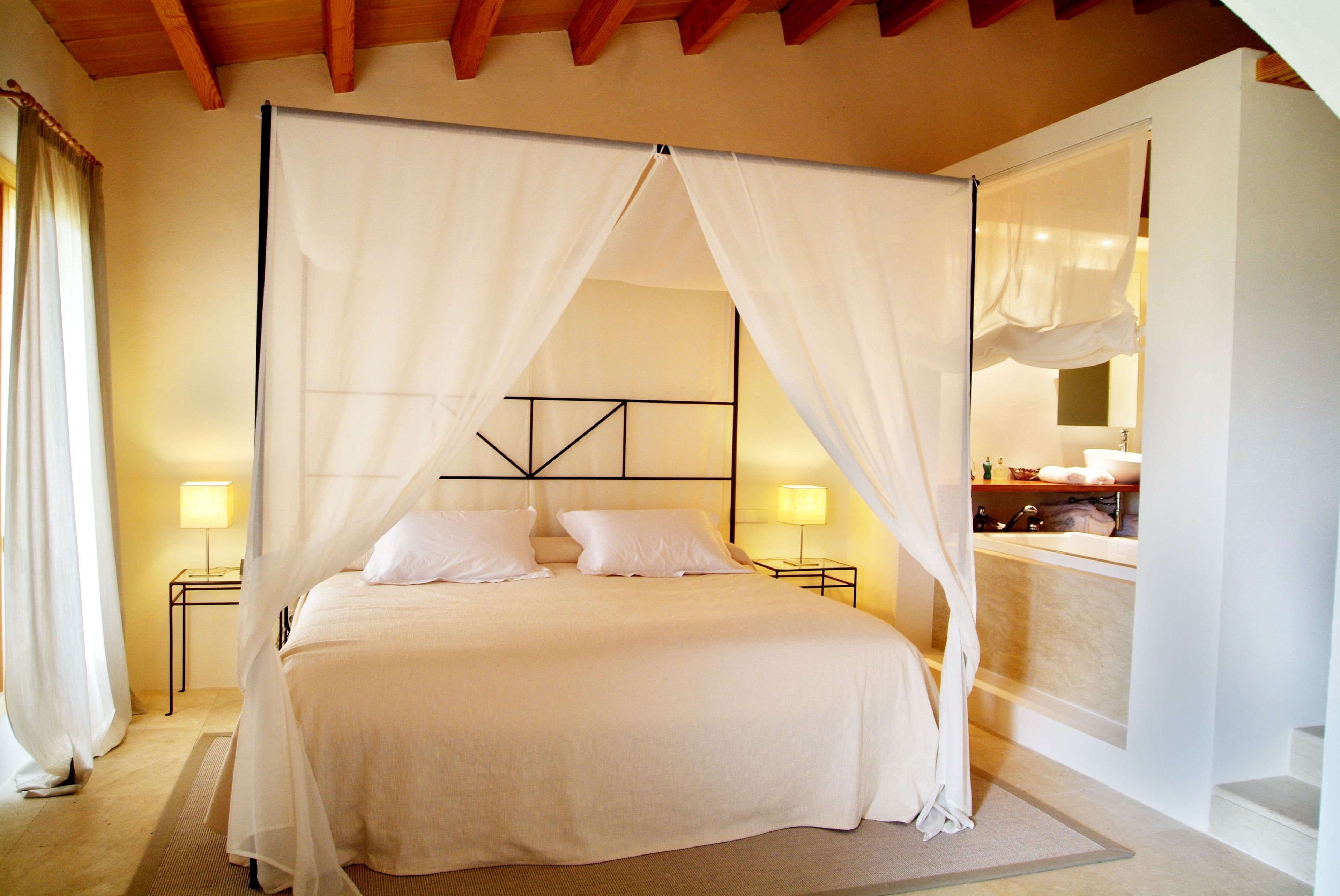 Quarto com cama de dossel, luminárias de cabeceira, mesa de apoio e banheiro visível através de uma porta aberta.