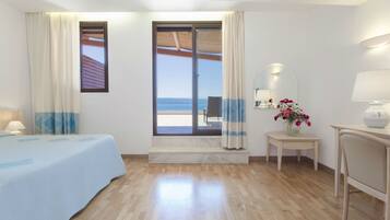 Quarto de hotel com cama, uma pequena mesa com abajur, espelho, um vaso com flores e vista para o mar através de uma porta deslizante.