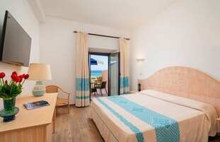 Quarto de hotel com cama, escrivaninha com luminária, cadeira, televisão e vista para o mar através de uma porta de vidro.