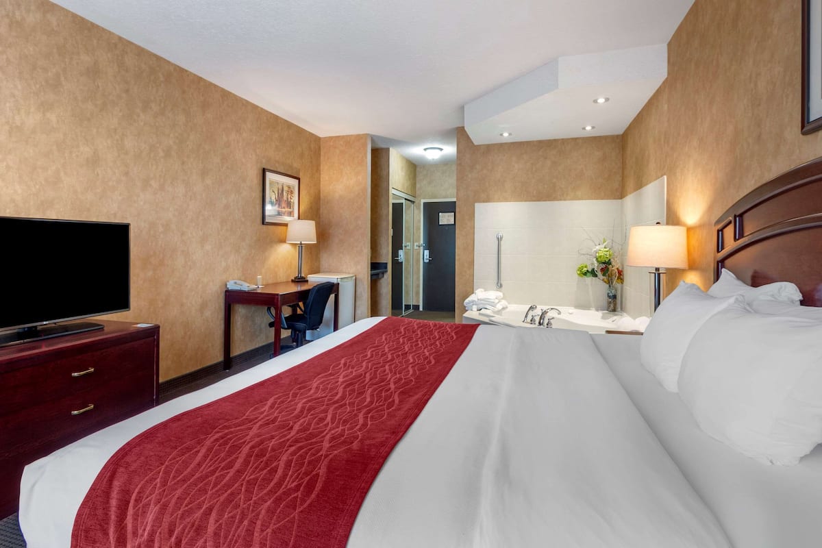 Quarto de hotel com uma cama grande, uma escrivaninha com cadeira, televisão, banheiro com espelho e porta que dá para outro cômodo.