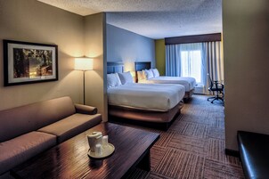 Quarto de hotel com sofá, mesa com bandeja e copos, duas camas, escrivaninha e uma cadeira.