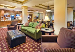 Um lobby de hotel com um sofá verde, um banquinho preto e uma poltrona estampada. Há um canto para café com uma placa que diz 'EXPRESS Start' e uma área de buffet com um refrigerador.