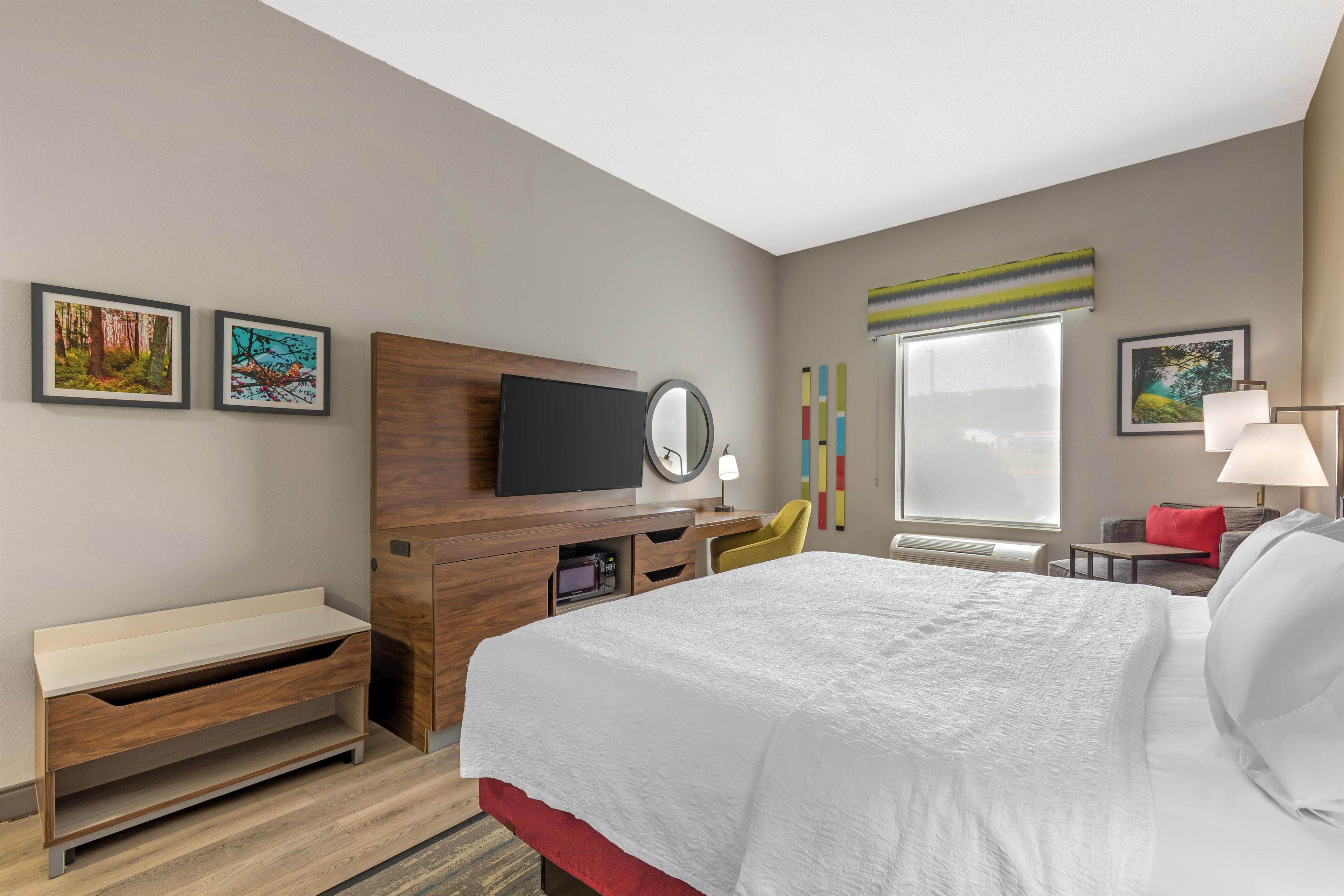 Quarto de hotel com cama, escrivaninha, cadeira, televisão e obras de arte nas paredes.
