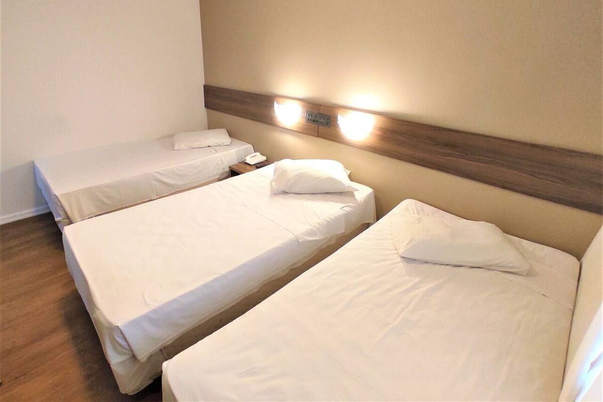 Quarto de hotel com duas camas de solteiro, uma cabeceira de madeira e um candeeiro fixado na parede.