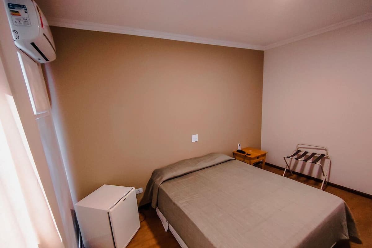 Um quarto pequeno e simples, com uma cama, uma mesinha, um radiador e um eletrodoméstico branco.