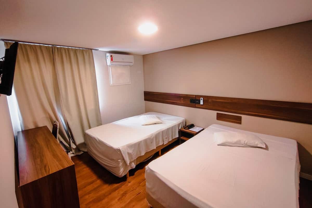 Quarto de hotel com duas camas de solteiro, uma escrivaninha de madeira, uma cadeira, televisão e janela com cortinas.