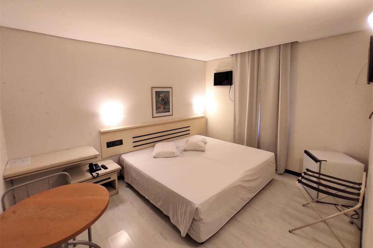 Quarto de hotel com cama, mesa de cabeceira, cadeira e uma pequena mesa.