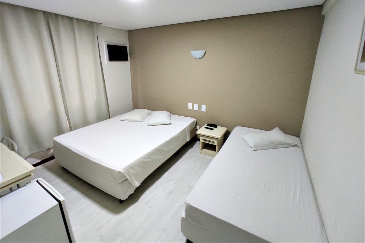 Quarto de hotel com duas camas, uma mesa de cabeceira com abajur, uma janela pequena e luminária na parede.