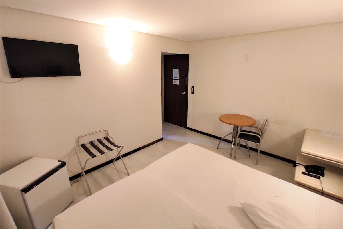 Quarto de hotel com cama, escrivaninha, cadeira, mesinha, televisão fixada na parede e porta.