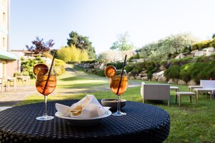 Duas bebidas sobre uma mesa com um prato de batatas fritas e uma tigela de nozes, em ambiente externo com vista para um jardim e um edifício ao fundo.