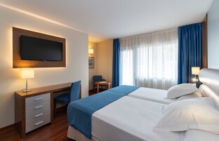 Quarto de hotel com televisão na parede, escrivaninha com gavetas, duas camas com roupa de cama branca, uma cadeira azul, uma mesa pequena e uma janela com cortinas azuis.
