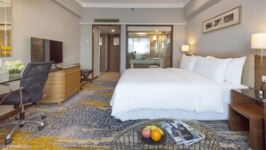 Quarto de hotel com uma cama grande, uma escrivaninha com cadeira, uma mesinha com fruta e uma revista, e um espelho na parede.