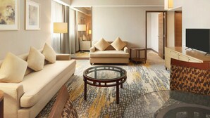 Quarto de hotel moderno com sofá, mesa de centro redonda de vidro, televisão e carpete estampado.
