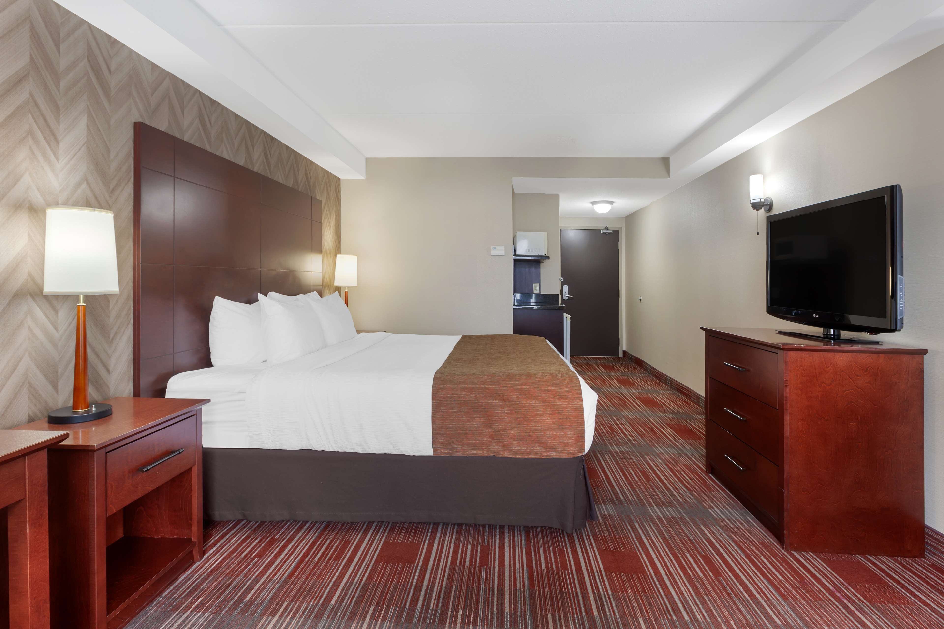Quarto de hotel com cama grande, televisão, secretária e cozinha compacta ao fundo.