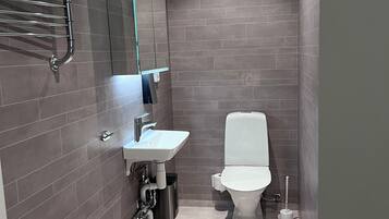 Um banheiro moderno com parede revestida de azulejos cinzas, pia branca, vaso sanitário e box de vidro.