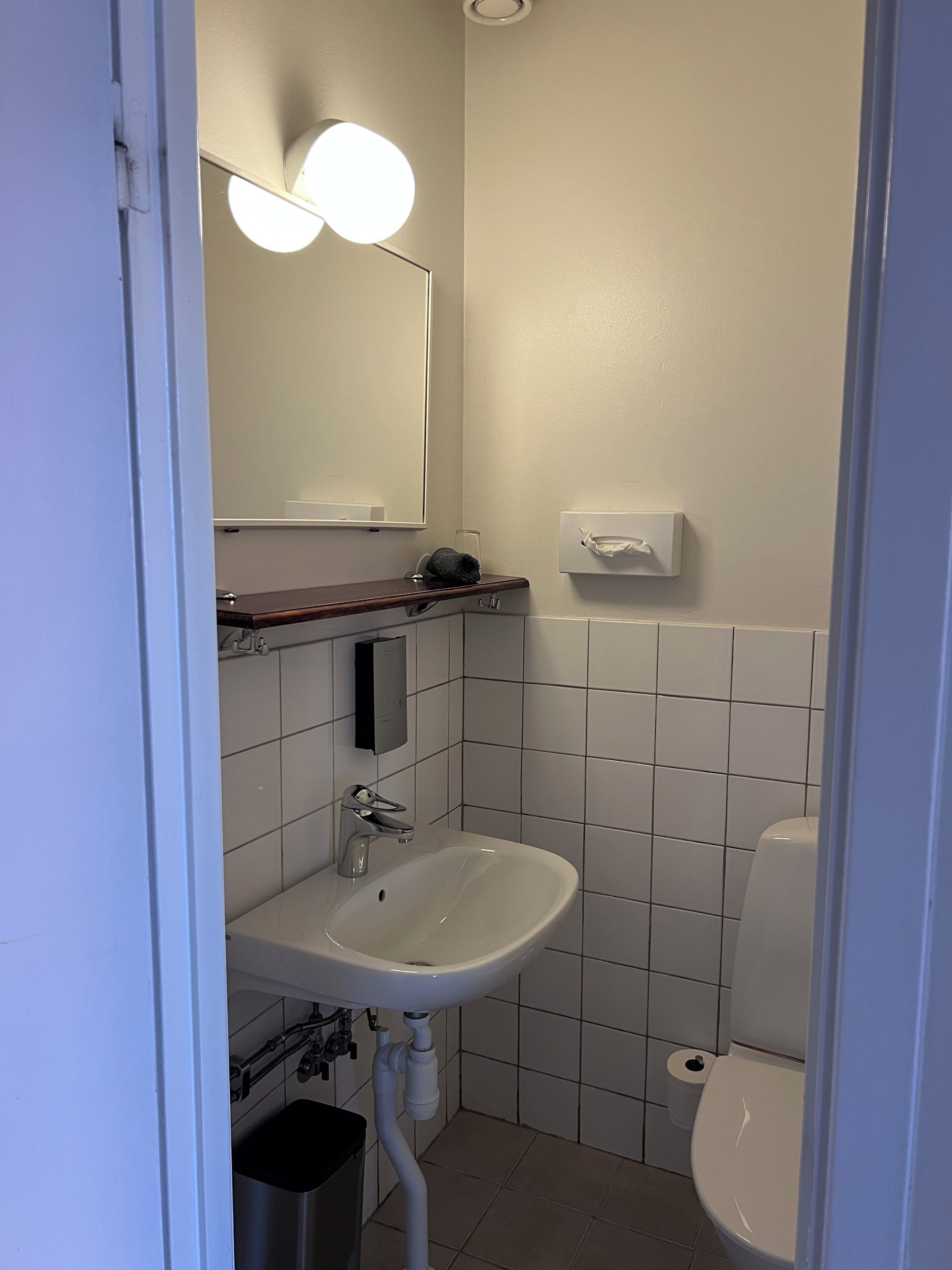 Casa de banho com lavatório branco, espelho e sanita. Dispenser de toalhas e dispenser de sabonete na parede.
