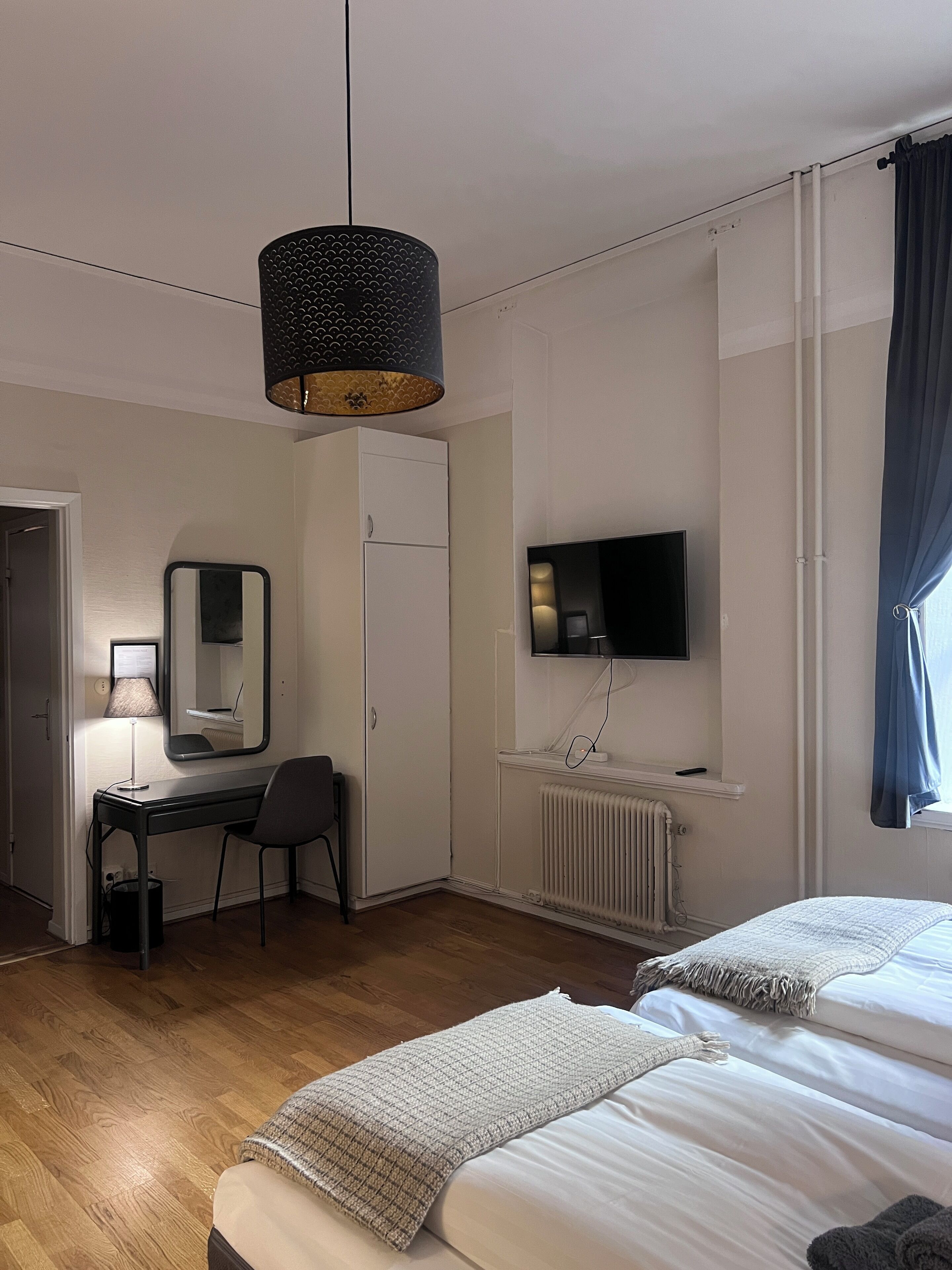 Quarto de hotel moderno com chão de madeira, uma escrivaninha com cadeira, uma cama com cobertor, televisão na parede e uma janela com cortinas.