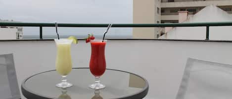 Uma mesa de vidro redonda com duas bebidas, guarda-corpo de varanda e um prédio ao fundo.