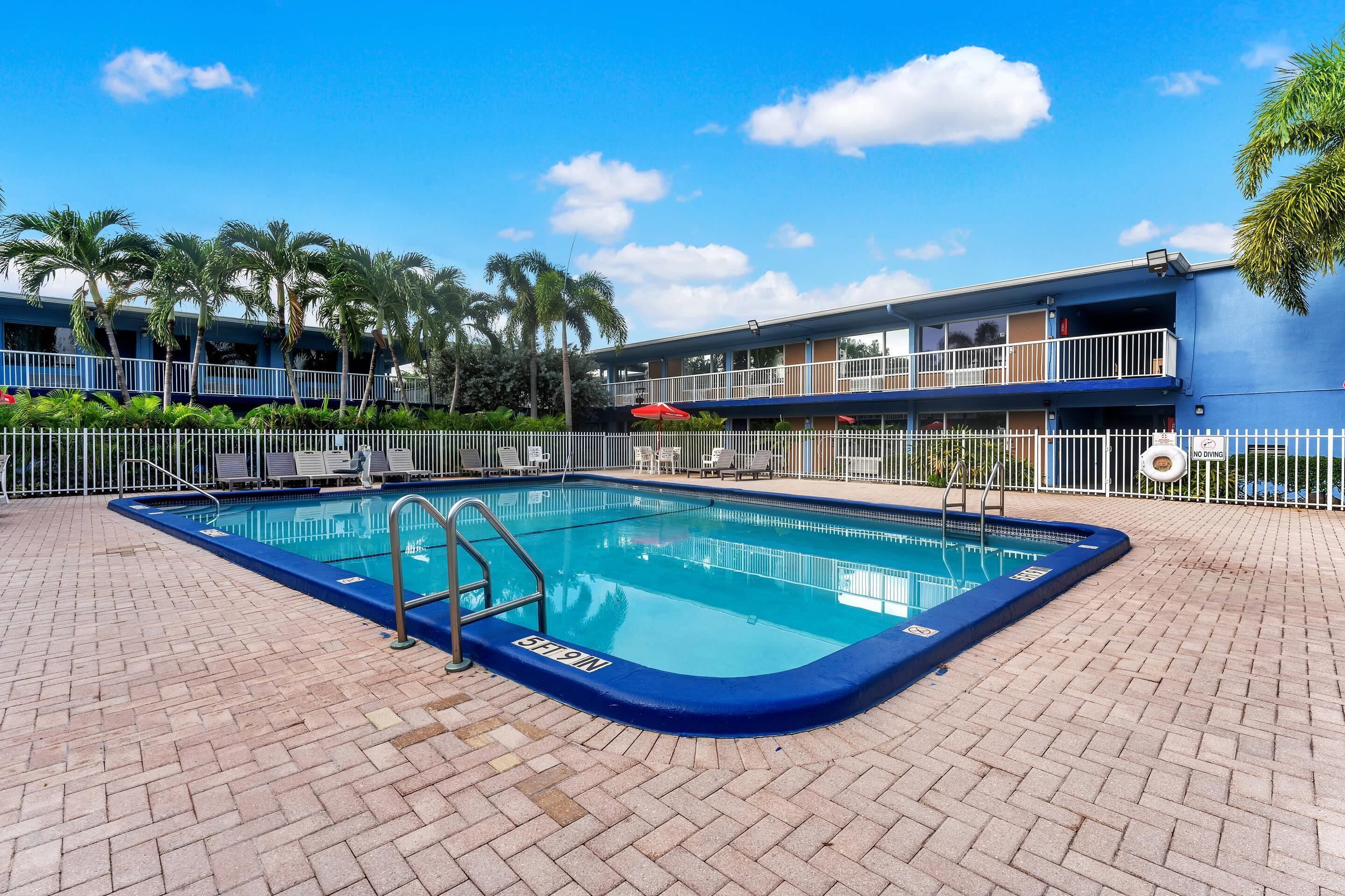 Piscina com borda azul, rodeada por uma cerca branca, e um edifício com varandas ao fundo.