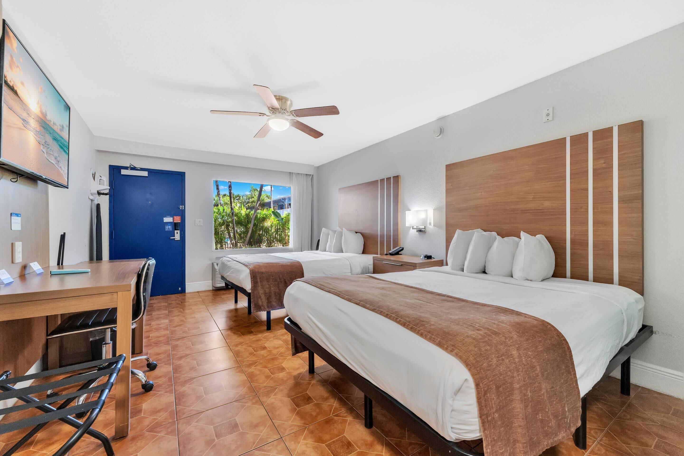 Quarto de hotel com duas camas, uma escrivaninha, uma cadeira, ventoinha no teto e vista para o verde lá fora.