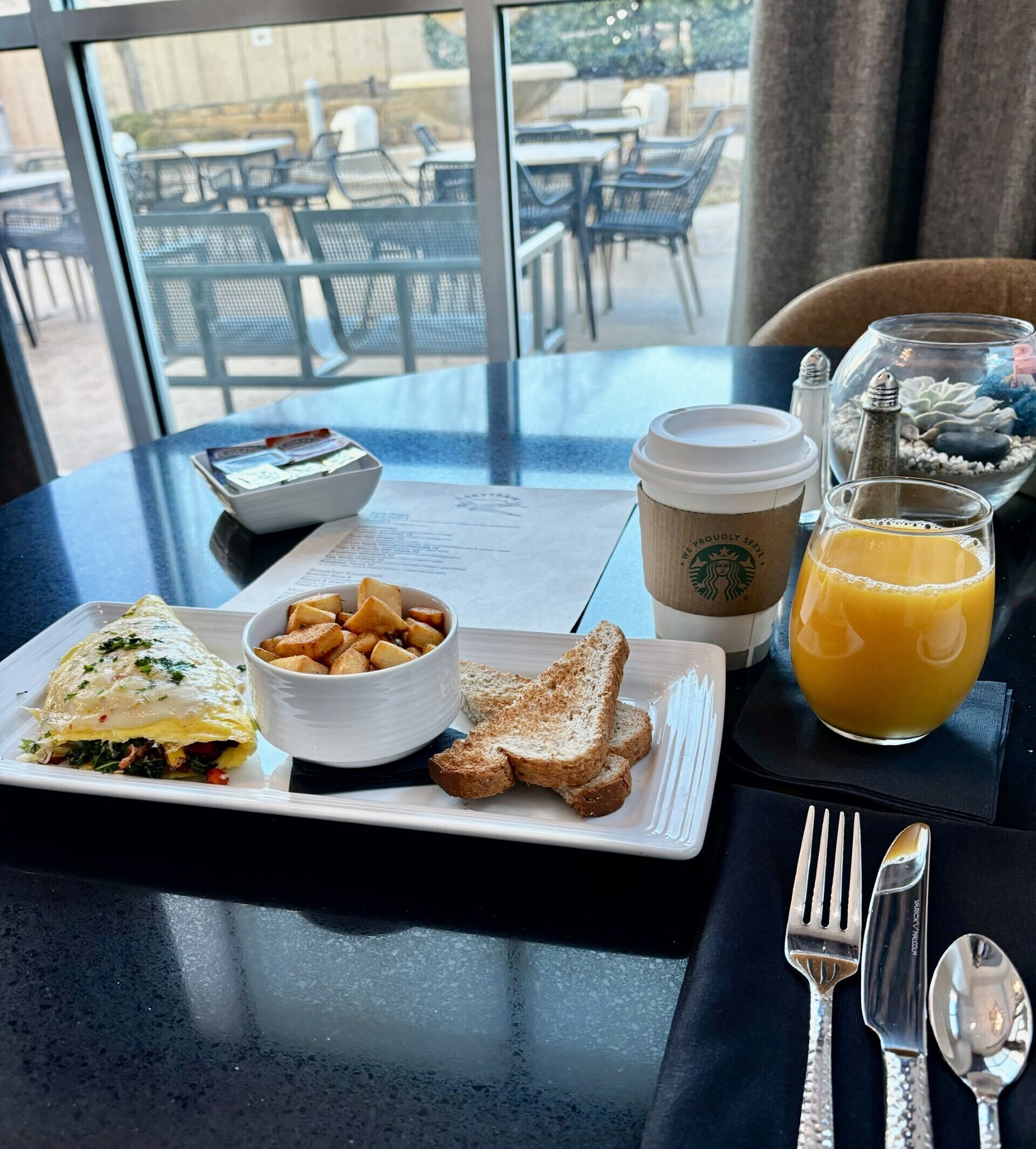 Bandeja de café da manhã com omelete, batatas, torradas, uma xícara da Starbucks e um copo de suco de laranja sobre uma mesa com vista para área externa.