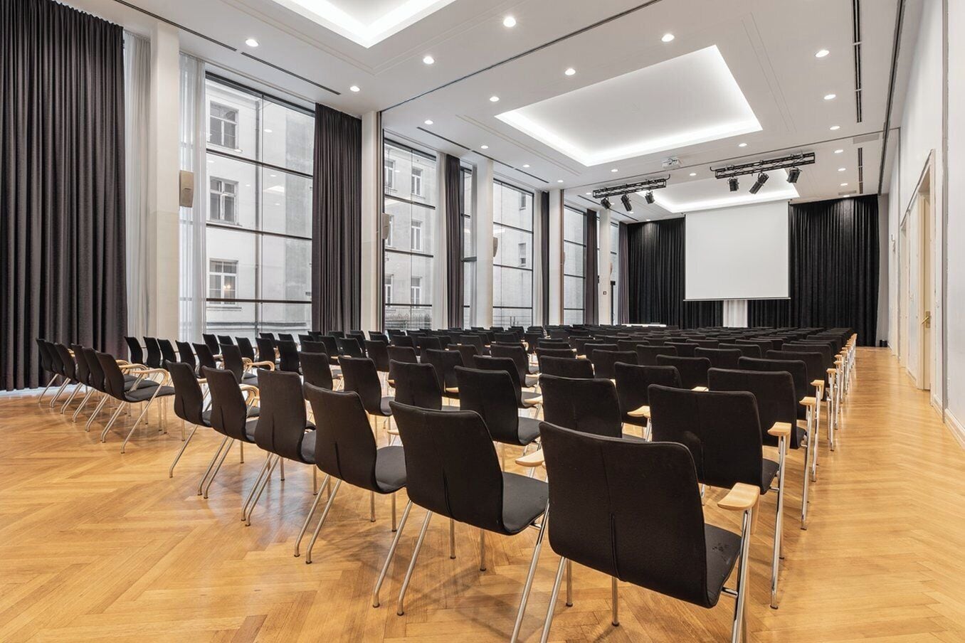 Uma sala de conferência com fileiras de cadeiras, janelas amplas e um palco com projetor.